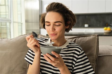 Retrato De Uma Jovem Mulher Morena Comendo No Sof Segurando Uma Tigela E Uma Colher Com Cereais