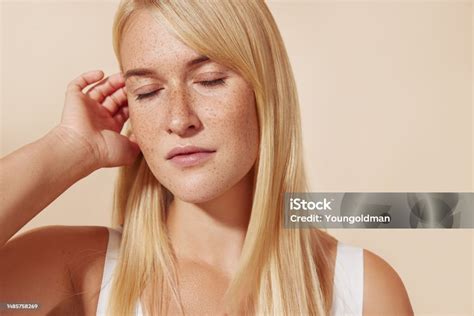 주근깨가 있는 피부를 가진 젊은 여성이 스튜디오에서 눈을 감고 서 있다 금발의 여자가 파스텔 톤을 배경으로 포즈를 취하면서 머리를 조정하고 있다 여자에 대한 스톡 사진 및