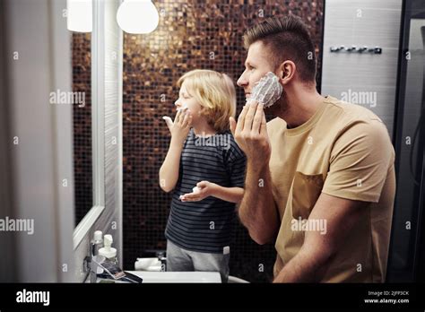 Side View Portrait Of Father And Son Shaving Together And Looking In