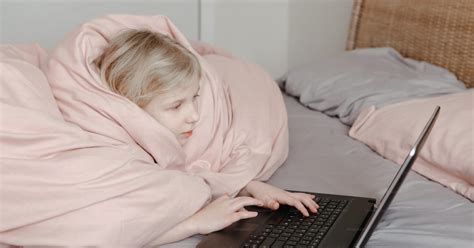 지루한 소녀는 담요 아래 침대에 누워 가상 온라인 학교 수업에서 학습합니다 집에서 노트북 인터넷으로 일하는 아이 무선 기술을 사용하는 어린이 교육 아이들을 위한 홈