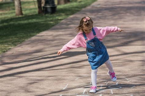 분홍색 옷을 입은 행복한 소녀는 거리의 아스팔트에 분필로 고전을 그립니다 화창한 여름날에 놀고 뛰어오르는 어린 소녀의 초상화 아이들의 창조적 발달 4 5세에 대한 스톡 사진