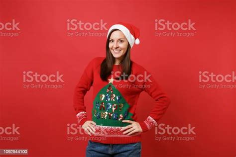 니트 스웨터 크리스마스 모자 Akimbo 밝은 빨간 벽 배경에 고립 된 무기와 서 웃는 젊은 산타 소녀 행복 한 새 해 2019 축 하 휴일 파티 개념입니다 복사 공간을