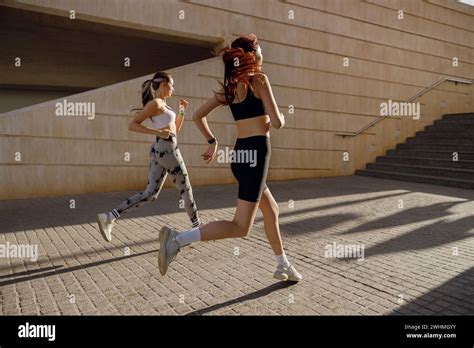Side View Of Women Athlete Running Side By Side Along An Outdoor Track On Buildings Background