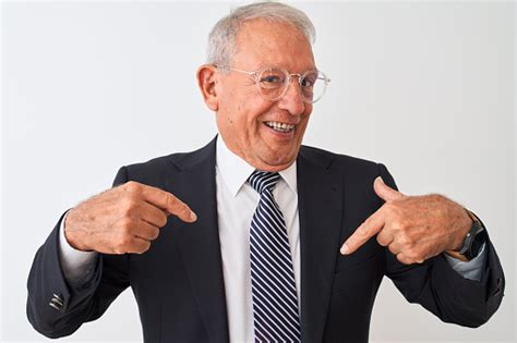 고립 된 흰색 배경 위에 양복과 안경을 착용한 선임 회색 머리의 사업가가 얼굴에 미소를 지으며 자랑스럽고 행복한 손가락으로 자신을 가리킵니다 가리키기에 대한 스톡 사진 및