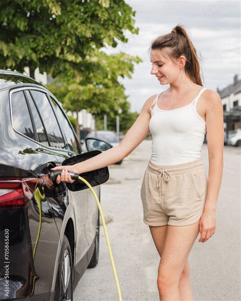 Girl In A Casual Summer Outfit Opening A Socket And Putting A Charger In A Black Electric Car