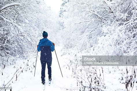 윈드브레이커와 모자를 쓰고 스키 폴을 손에 들고 눈 덮인 숲을 배경으로 등을 대고 모자를 쓰고 있는 스키어 겨울 숲에서 크로스 컨트리 스키 야외 스포츠 건강한 라이프 스타일