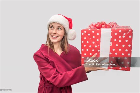 행복 하 고 궁금해 하는 젊은 여성이 서 서 왼쪽으로 보인다 그녀는 빨간 코트와 크리스마스 모자를 착용 합니다 모델은 손에 큰 존재를 보유 하 고 있습니다 그녀는 흥분 된다