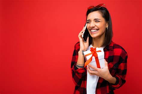 매력적인 즐거운 감정적 인 성인 갈색 머리 여자는 빨간색 리본흰색 선물 상자를 들고 흰색 캐주얼 티셔츠와 빨간색과 검은 셔츠를 입고 빨간색 배경 벽에 고립 하고 휴대 전화에