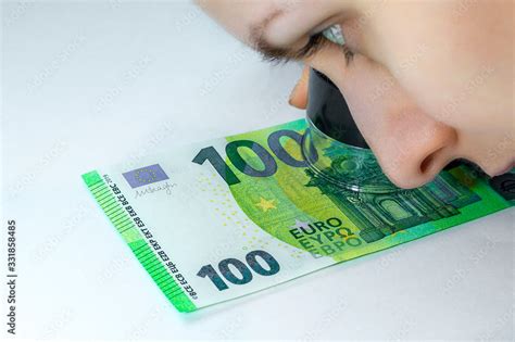 The Girl Employee Of The Bank Checks The Banknote For Counterfeit Using A Special Magnifier