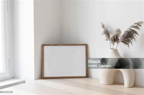 마른 풀 현대 세라믹 꽃병에 갈대 식물 꽃다발 창문 근처의 나무 테이블에 흰 벽에 빈 사진 프레임 모형 미니멀리즘 스칸디나비아 스타일 우아한 보헤미안 인테리어 거실 디자인에