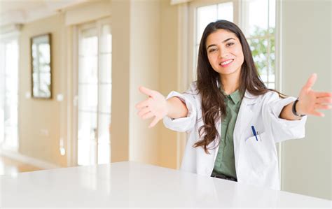 치료사 나 의사로 병원에서 의료 코트를 입고 젊은 여성은 포옹을 위해 열린 팔로 웃 고 카메라를 보고 행복을 품은 쾌활 한 표현