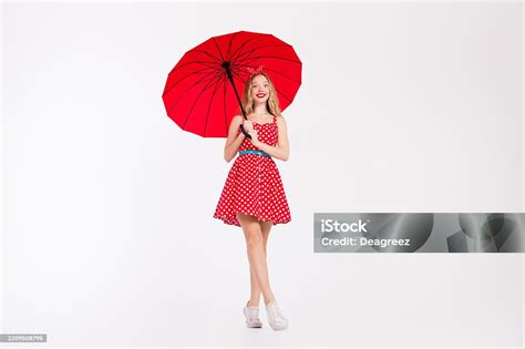 빈티지 물방울 무늬 드레스를 입은 장난기 많은 여성이 흰색 배경에 빨간 우산을 쓰고 포즈를 취하며 복고풍 매력과 활기찬 매력을