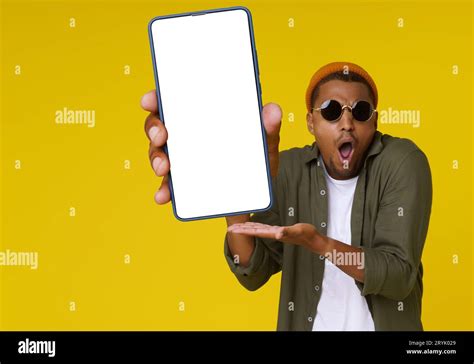 surprised african man is holding phone with white blank screen against yellow background with