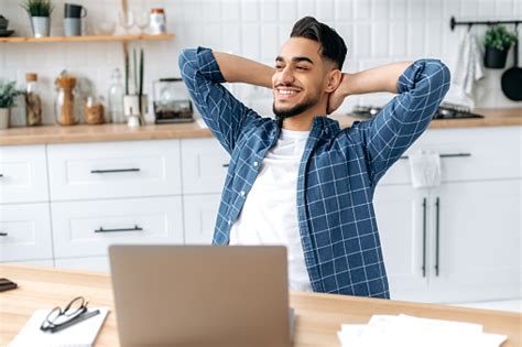 행복한 아라비안 또는 인도 사람 프리랜서 집에서 일하고 직장에서 부엌에 앉아 직장에서 휴식을 취하고 머리 뒤에 손을 얹고 옆을 바라보고 휴식을 꿈꾸고 미소 짓는다 재택근무에