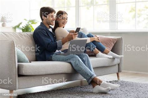 기쁜 쾌활한 밀레니엄 세대 유럽 남자와 여자 노트북과 소파에 앉아 스마트 폰을보고 2명에 대한 스톡 사진 및 기타 이미지 2