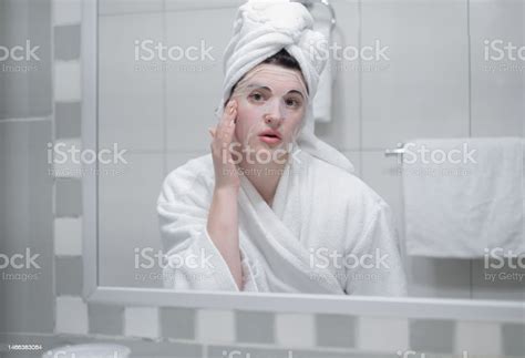 흰색 목욕 가운을 입고 머리에 수건을 두른 젊은 백인 여성이 얼굴에서 보습 시트 마스크를 제거하고 욕실에서 뷰티 트리트먼트를 합니다 25 29세에 대한 스톡 사진 및 기타