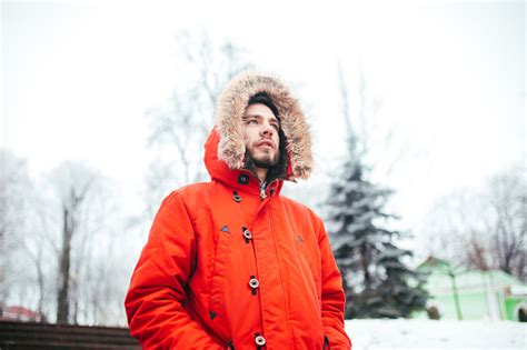 빨간색 겨울 자 켓 후드와 그의 머리에 모피를 입은 수염과 세련 된 젊은이 눈 덮인 시의 배경으로 서 하 고 사려깊은 보인다 겨울와 서 리 테마 겨울에 대한 스톡 사진 및