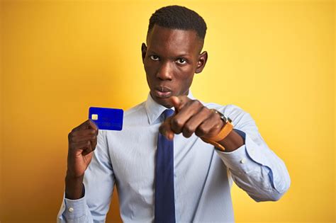 카메라와 당신에 게 손가락으로 가리키는 고립 된 노란색 배경 위에 서 신용 카드를 들고 아프리카 계 미국인 사업가 손 기호 앞에서 긍정적이 고 자신감 제스처 손에 대한 스톡