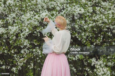 꽃이 만발한 벚나무 배경에 흰 블라우스를 입은 금발 소녀 짧은 분홍색 머리 스커트를 입은 모델 깃털로 뷰티 메이크업 스타일과 패션 오뜨 꾸뛰르 여자에 대한 스톡 사진 및
