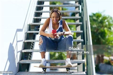 색깔이 있는 머리띠를 가진 흑인 여성 스케이트보드에 발을 얹고 스마트폰을 상담한다 20 24세에 대한 스톡 사진 및 기타 이미지 20 24세 Brand Name