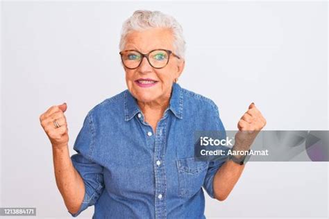 데님 셔츠와 안경을 착용한 선임 회색 머리의 여성은 매우 행복하고 팔을 들고 미소를 지으며 성공을 위해 비명을 지르는 우승자 제스처를하고 흥분했습니다 축 하 개념입니다