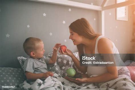 임산부와 어린 소년 아들은 아침에 집에 있는 침대에서 사과와 복숭아를 먹고 있습니다 침실에서 캐주얼 라이프 스타일 가정 생활에
