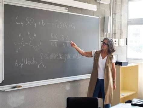 Female Teacher Explaining Mathematical Or Physics And Chemistry Formulas On A Blackboard In A