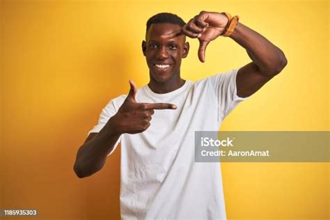 행복한 얼굴로 손과 손가락으로 프레임을 만드는 고립 된 노란색 배경 위에 서있는 흰색 티셔츠를 입은 젊은 아프리카 계 미국인 남자 창의력과 사진 개념 남자에 대한 스톡 사진