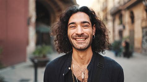 긴 곱슬 머리와 그루터기를 가진 젊은 매력적인 이탈리아 남자는 카메라를 들여다 보며 오래된 건물 배경을 미소 짓습니다 귀에