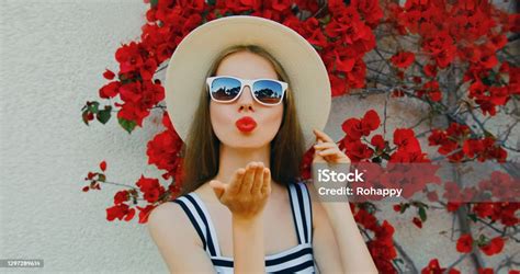 빨간 꽃 배경에 여름 밀짚 모자를 쓰고 달콤한 공기 키스를 보내는 빨간 입술을 불고있는 아름다운 여성의 초상화 20 29세에 대한 스톡 사진 및 기타 이미지 Istock