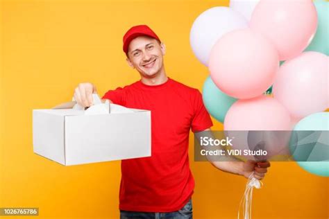 음식 주문 케이크 상자 노란색 배경에 절연을 주는 남자 남성 직원 택배 빨간 모자 티셔츠 보류 다채로운 공기 풍선에 빈 골 판지 상자에서 디저트 배달 서비스 개념입니다