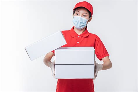 빨간 모자 빈 티셔츠 유니폼 얼굴 마스크 장갑을 낀 의식이 있는 젊은 배달원의 이미지는 밝은 회색 배경 스튜디오에 격리된 빈 흰색 판지 상자 팬데믹 코로나바이러스 바이러스