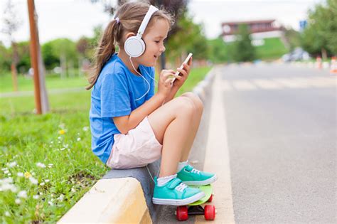 화창한 여름 날에 공원에서 스케이트 보드와 연석에 작은 귀여운 소녀와 흰색 헤드폰의 음악을 듣고 라이프 스타일 행복한 여름 여름 야외 초상화 귀여운에 대한 스톡 사진 및