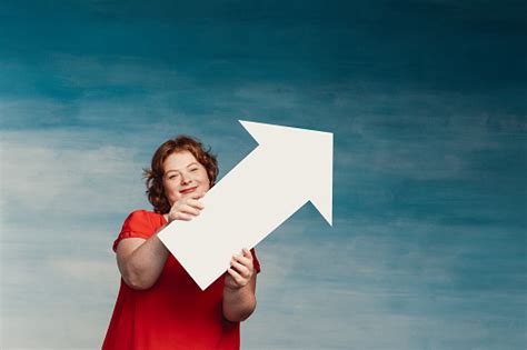 빨간 티셔츠와 청바지에 예쁜 빨간 머리 통통 한 여자는 파란색 배경에 측면에 흰색 위로 화살표를 보유하고 있습니다 T 셔츠에