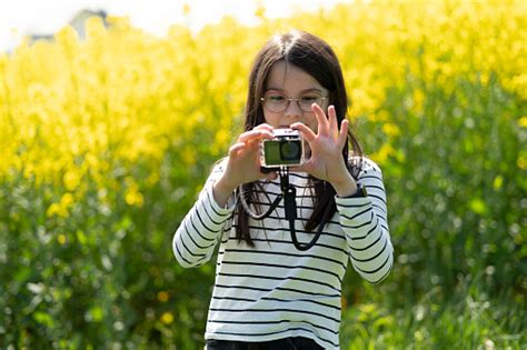 긴 머리에 안경을 쓰고 줄무늬 티셔츠를 입은 아름다운 소녀가 봄에 노란 유채밭 앞에 서서 카메라에 사진을 찍고 있습니다 어린이의 창의력과 여가의 개념 10 11세에 대한