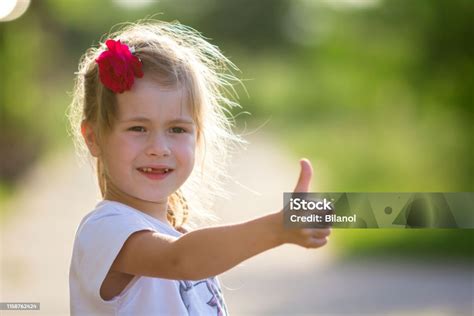 흐릿한 밝은 녹색 여름 배경에 카메라에 엄지 손가락 제스처를 보여주는 머리에 빨간 장미와 흰색 티셔츠에 귀여운 작은 금발 심각한