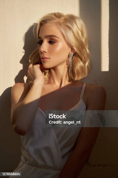 흰색 드레스를 입은 금발의 미녀 여성이 저녁 햇살의 창문에 서 있습니다 야생 꽃 자연 메이크업의 꽃다발을 가진 소녀의 초상화 아름다움에 대한 스톡 사진 및 기타 이미지