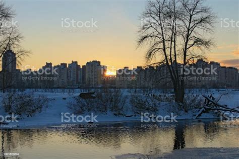 일몰 동안 얼어 붙은 드니퍼 강의 그림 같은 겨울 풍경 배경에 오볼론 지역의 건물 물에 반사 된 벌거 벗은 나무 태양이 집 뒤에 숨어 있습니다 키예프 0명에 대한 스톡 사진