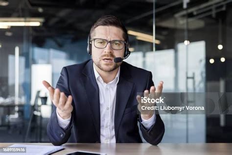 헤드셋을 착용한 피곤하고 속상한 청년이 카메라 앞 사무실에 앉아 화상 통화로 불행하게 이야기하는 초상화 손으로 올리고 몸짓 서비스에 대한 스톡 사진 및 기타 이미지 Istock