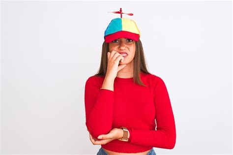 고립 된 흰색 배경 위에 서있는 프로펠러와 패니 캡을 입고 젊은 아름다운 소녀는 입물고 손톱에 손을 긴장 찾고 스트레스와 긴장 불안 문제 갈색 머리에 대한 스톡 사진 및