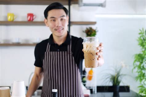 커피숍카운터바에서 아이스 라떼 커피 한 잔을 마시며 미소짓는 아시아 바리스타 청년 투명 플라스틱 유리를 들고 손에 초점을 닫습니다 명랑한에 대한 스톡 사진 및 기타 이미지