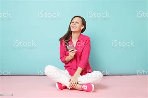 전체 길이 세로 여자 장미 셔츠 블라우스 이어폰으로 바닥에 앉아 흰 바지에에서 밝은 핑크 블루 파스텔 벽 배경 스튜디오에 고립 패션 라이프 스타일 개념입니다 복사 공간을