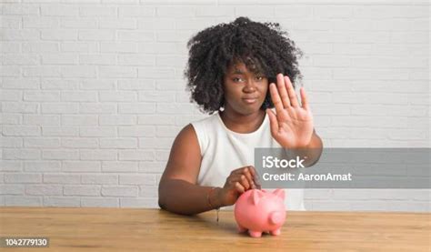 젊은 아프리카계 미국인 여자와 함께 돼지 저금통을 들고 테이블에 앉아 열 심각 하 고 자신감 표현 방어 제스처 정지 표시를 하 고 손을 경계 표지에 대한 스톡 사진 및 기타