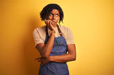 고립 된 노란색 배경 위에 앞치마를 입고 공포를 가진 아프로 미국 바리스타 남자는 손톱을 물고 손톱을 물고 손에 스트레스와 긴장 찾고 불안 문제 감정에 대한 스톡 사진 및