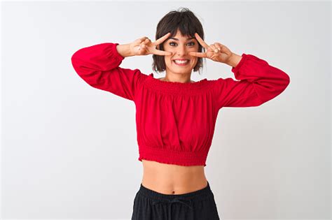 고립 된 흰색 배경 위에 서있는 빨간 여름 티셔츠를 입은 젊은 아름다운 여성은 얼굴에 손가락으로 평화 의 상징을하고 승리를 보여주는 쾌활한 미소 T 셔츠에 대한 스톡 사진