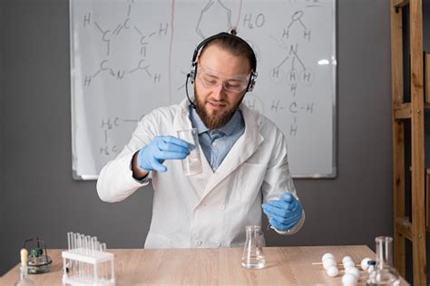 하얀 옷을 입은 화학 교사가 공식이 적힌 마커 보드를 배경으로 의자에 앉아 손에 액체가 든 플라스크를 들고 있습니다 웹캠 보기 기치 학습에 대한 스톡 사진 및 기타 이미지