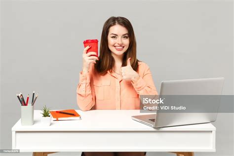커피 또는 차의 종이 컵을 들고 엄지 손가락을 보여주는 젊은 쾌활 한 여자는 회색 배경에 고립 된 Pc 노트북 책상에서 작업을 앉아 성취 비즈니스 커리어 라이프 스타일 컨셉
