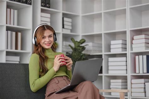 Smiling Asian Woman Using Webcam While Wearing Headphones And Sitting On Sofa At Home Stock