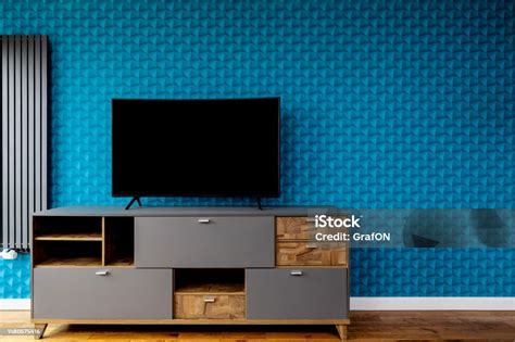 파란색 무늬 벽지가 있는 벽입니다 현대적인 회색 라디에이터 서랍이 있는 서랍장과 대형 평면 Tv가 있습니다 많은 장식 라디에이터 커튼 촛불 램프 촛대 냄비에 녹색 식물 0