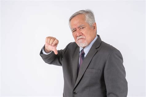 정장과 넥타이를 착용한 고위 아시아 사업가가 단호한 표정으로 불만을 표하고 엄지손가락을 내리는 제스처를 취하며 평범한 흰색 배경에 서 있습니다 70 79세에 대한 스톡 사진